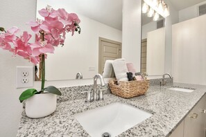 Casa de banho com bancada de granito, duas pias, um espelho, uma orquídea em vaso e uma cesta com toalhas.