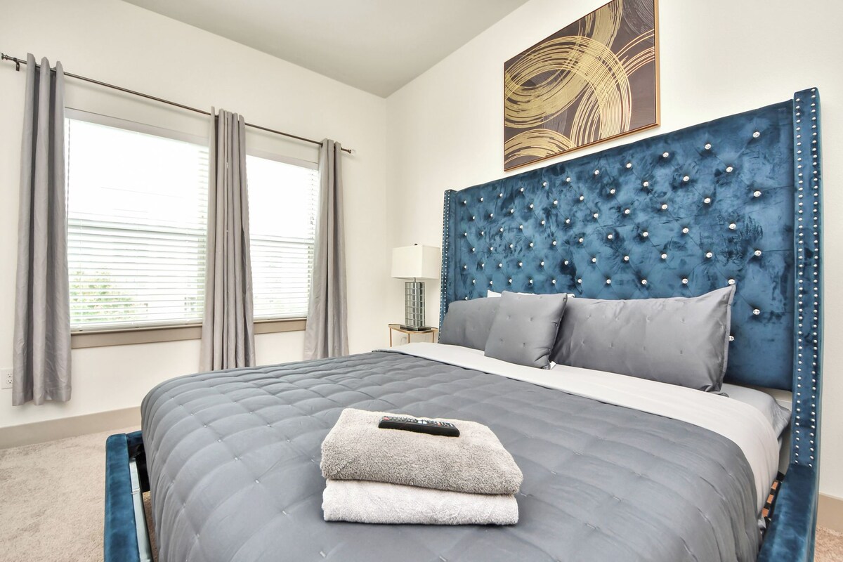 Quarto com cama grande, cabeceira com botões, controle remoto e janela com cortinas.