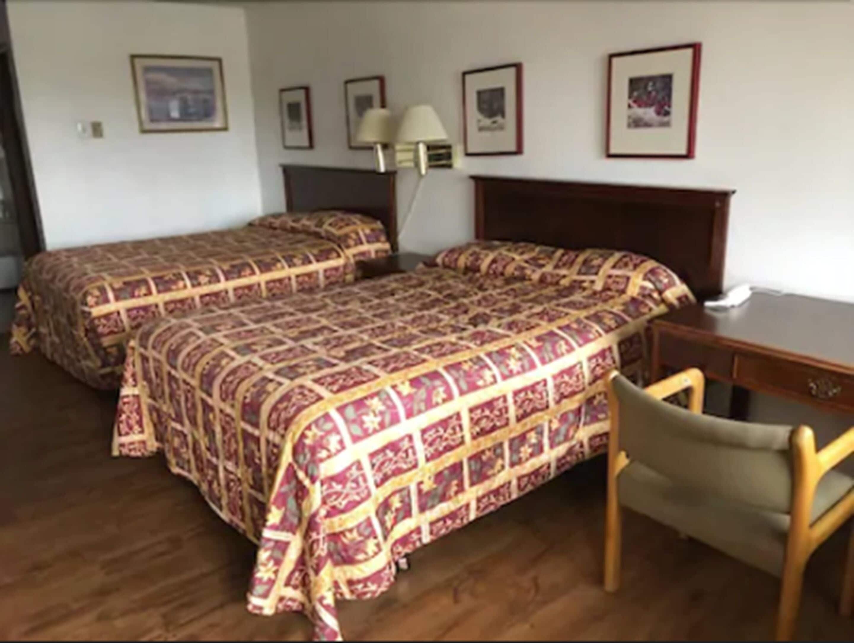 Quarto de hotel com duas camas, uma escrivaninha e uma cadeira. Há quadros nas paredes.