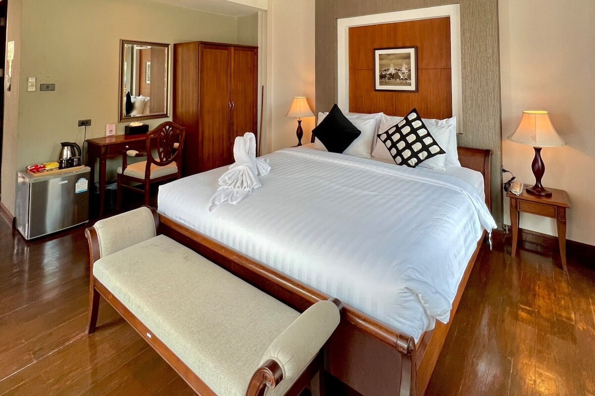 Quarto de hotel com uma cama grande, cabeceira de madeira, duas mesas de cabeceira com abajures, uma escrivaninha com cadeira, um frigorífico pequeno e um banco.