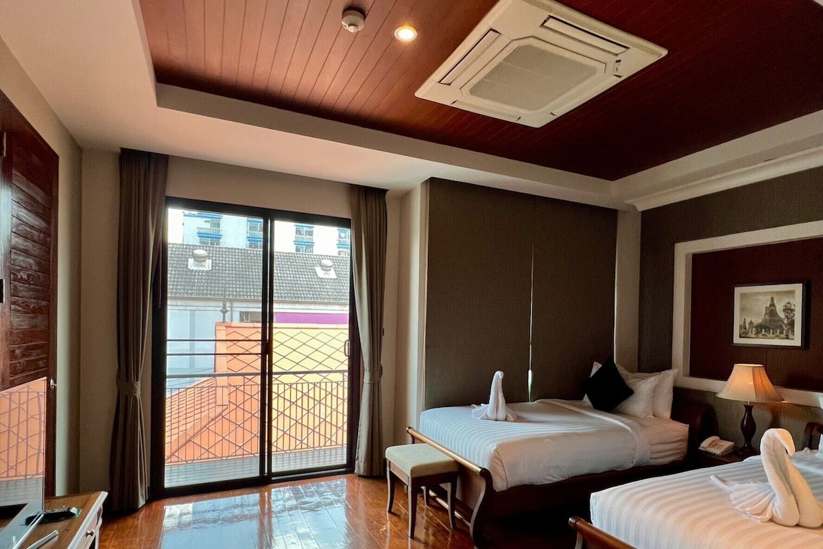 Quarto de hotel com duas camas, uma escrivaninha, um banco, porta de vidro deslizante para a varanda e um respiradouro no teto.
