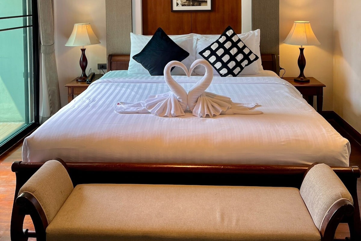 Quarto de hotel com uma cama grande, duas luminárias de cabeceira, um quadro na cabeceira e um banco no pé da cama.