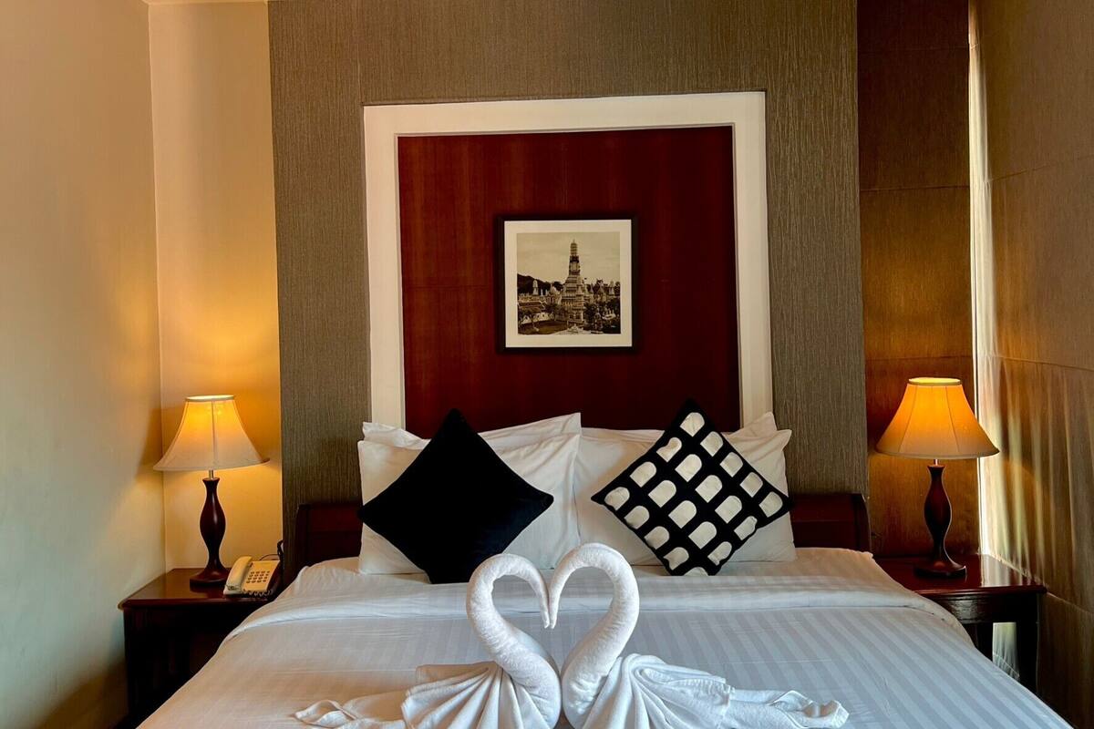 Quarto de hotel com cama grande, duas luminárias de cabeceira, um quadro na cabeceira e toalhas dispostas em formato de coração na cama.