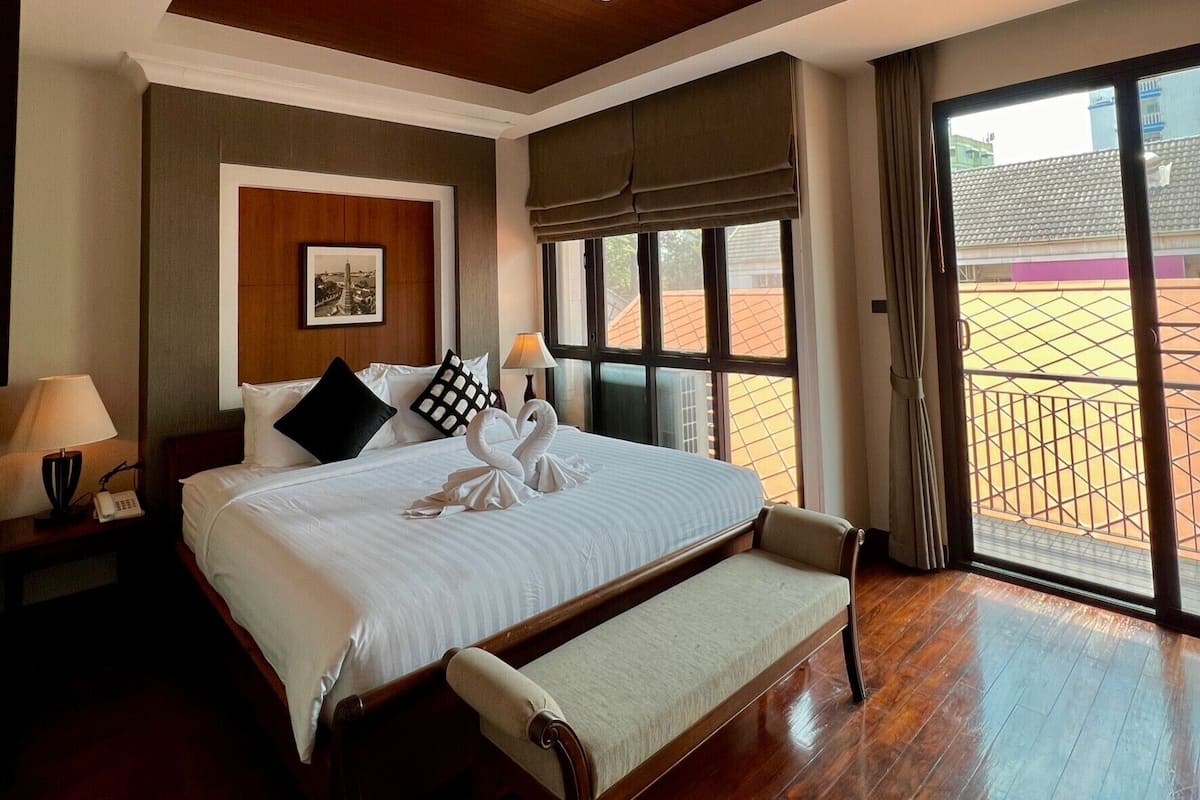 Quarto de hotel com uma cama grande, um banco, uma mesa de cabeceira com abajur e vista para o exterior através de uma janela ampla.
