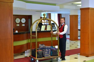 Uma área de recepção de hotel com um balcão de madeira, dois relógios e um carrinho de bagagem com malas.