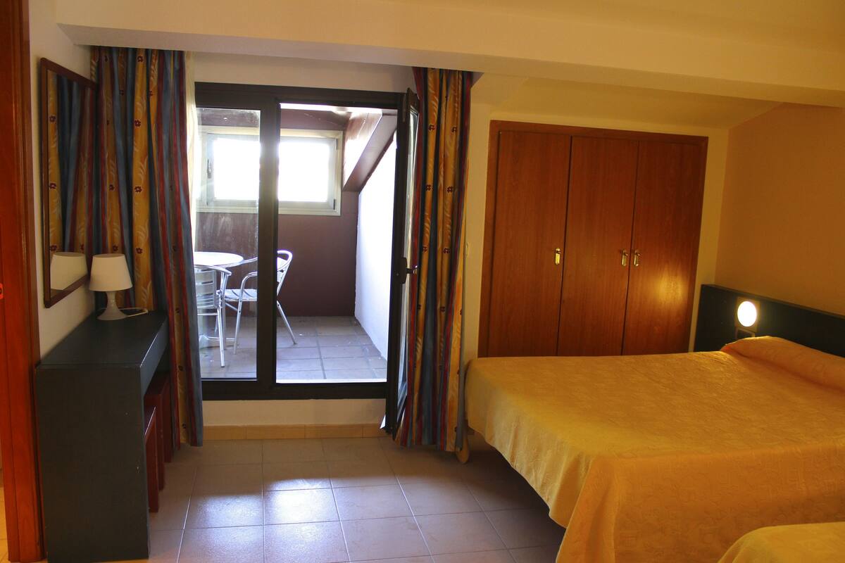 Quarto de hotel com uma cama, uma escrivaninha com candeeiro, um guarda-roupa e vista para área externa com mesa e cadeiras.