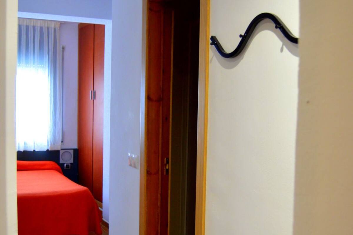 Um corredor leva a um quarto com colcha vermelha, porta de madeira e um gancho preto fixado na parede.