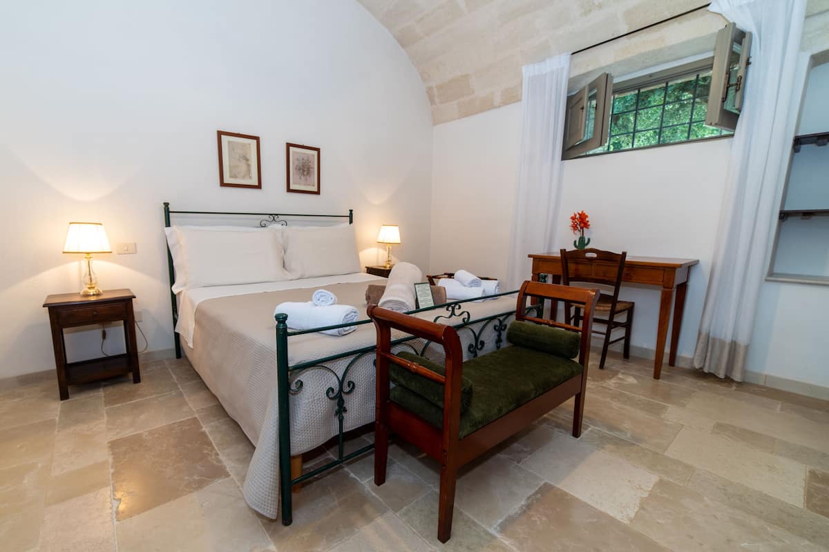 Quarto com cama, mesas de cabeceira, cadeira de madeira, escrivaninha e janela com cortinas.