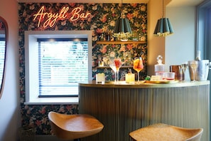 Uma área de bar com papel de parede floral, um letreiro de neon com a inscrição 'Argyll Bar' e uma janela com persianas. Há dois bancos de bar e uma variedade de bebidas no balcão.