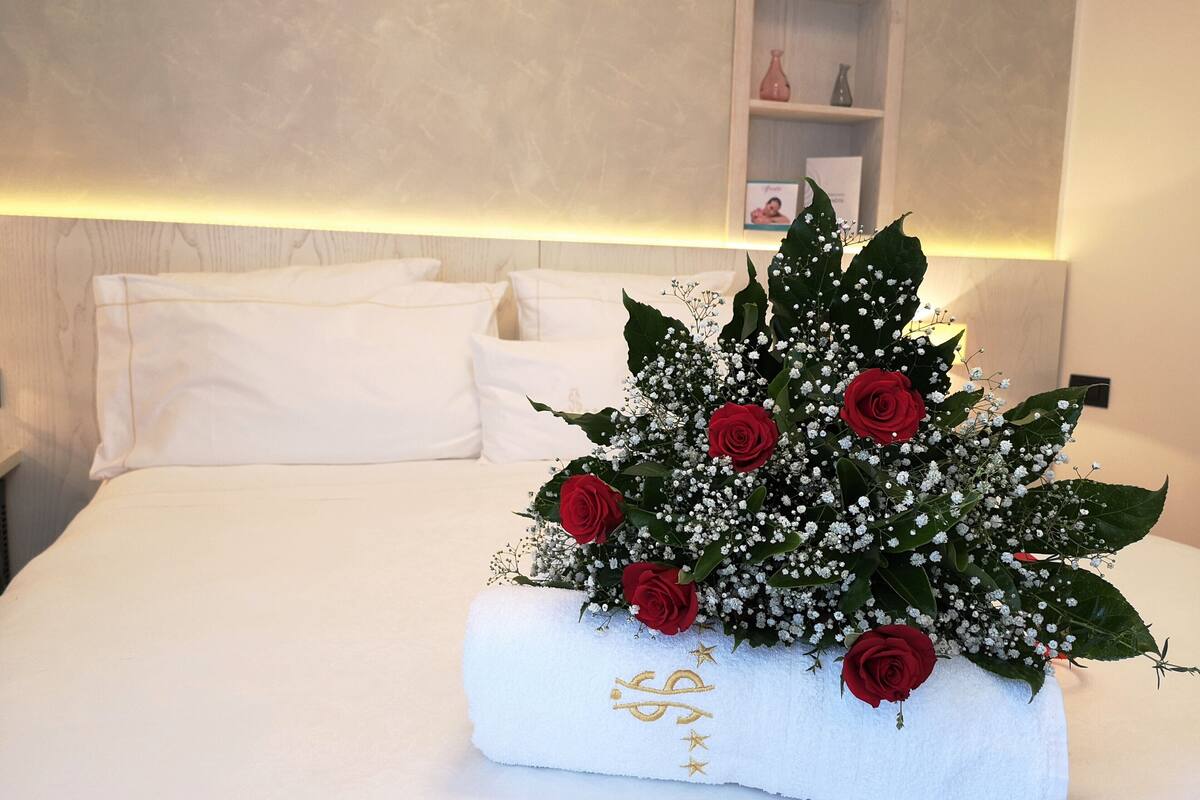 Cama bem arrumada com lençóis brancos e um arranjo floral com rosas vermelhas e respingos de mosquitinho.