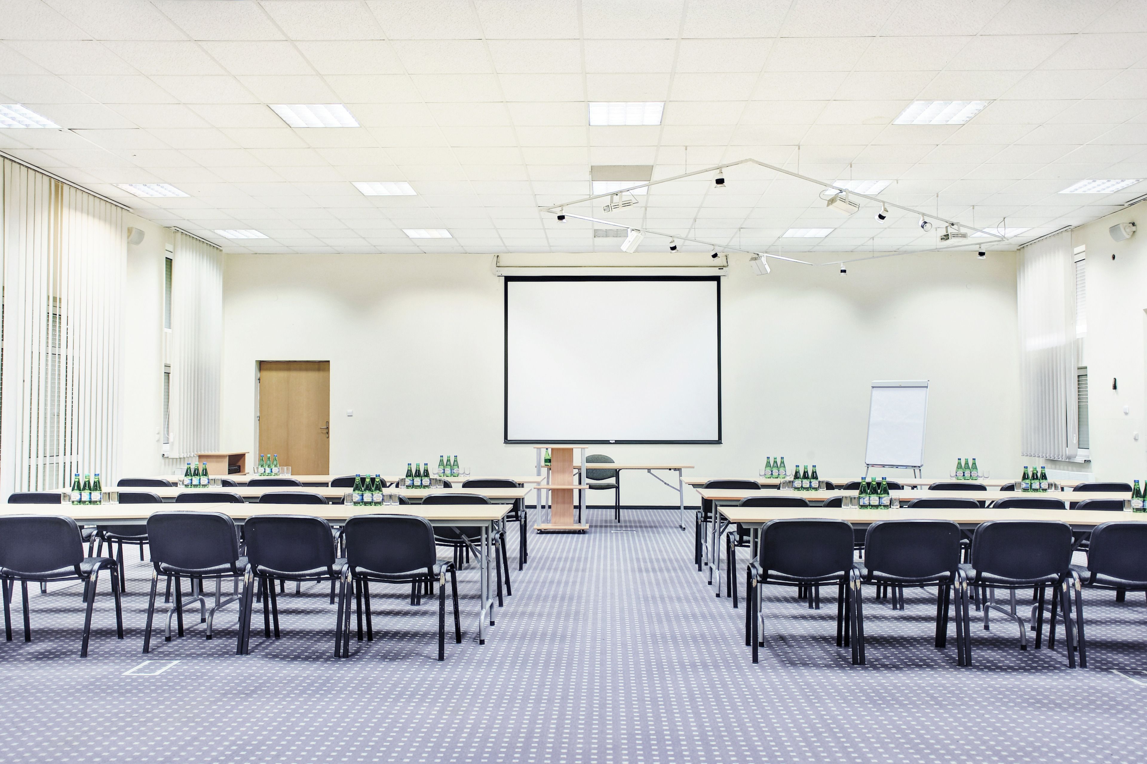 Sala de conferências com uma tela grande, fileiras de mesas com cadeiras e garrafas de água.