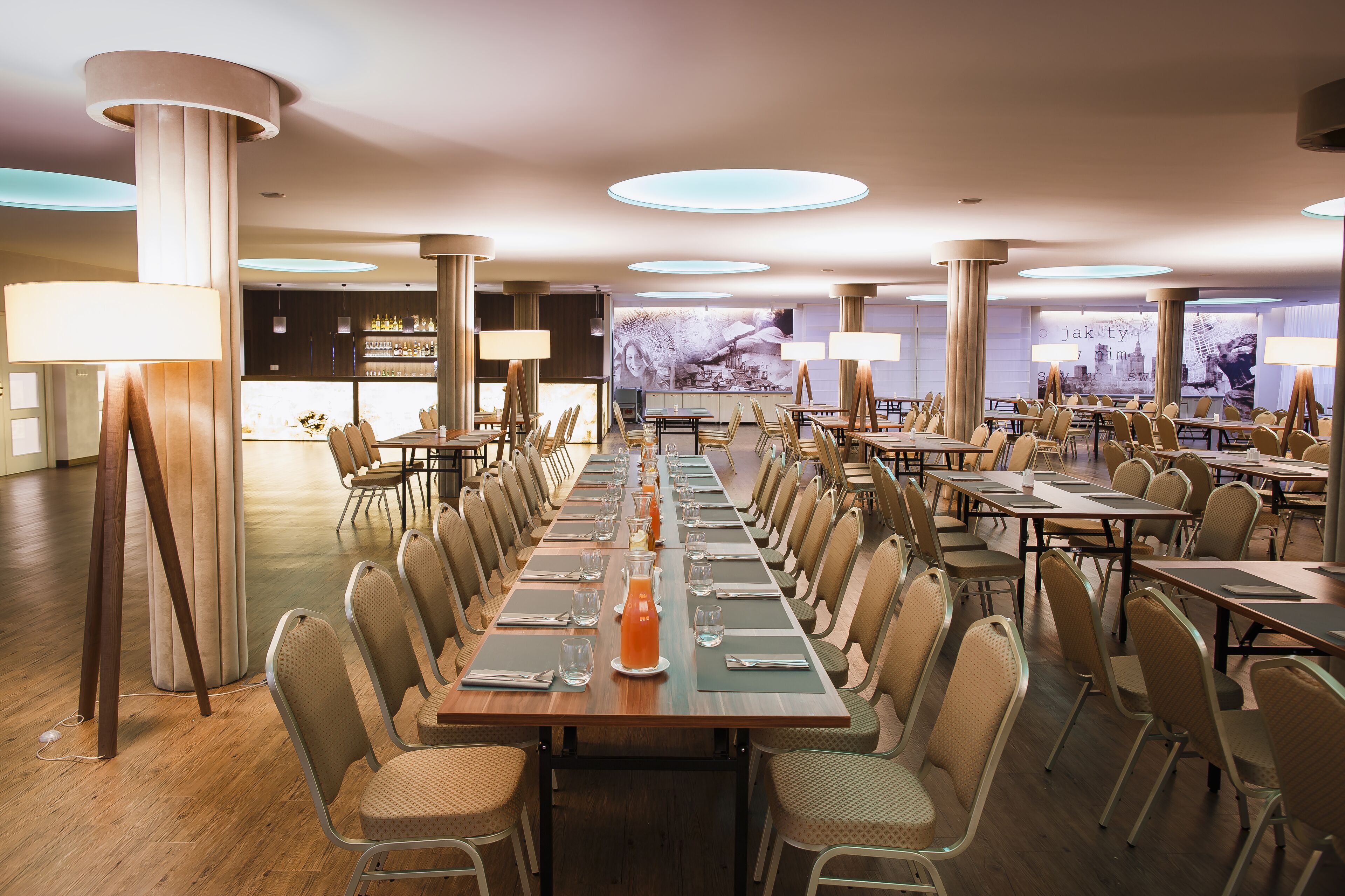 Uma sala de conferências ampla, com mesas compridas, cadeiras e um espaço de bar ao fundo.