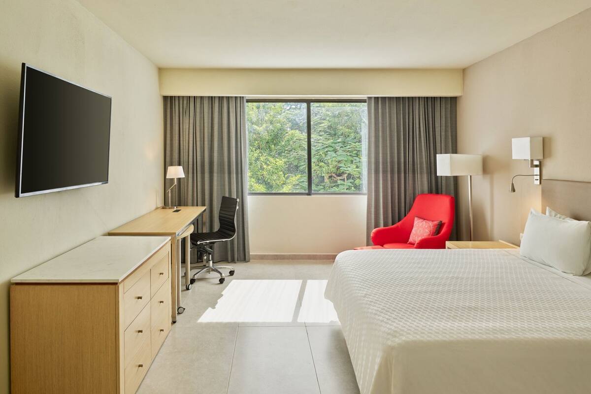 Quarto de hotel com cama, escrivaninha, cadeira, televisão e janela com cortinas.