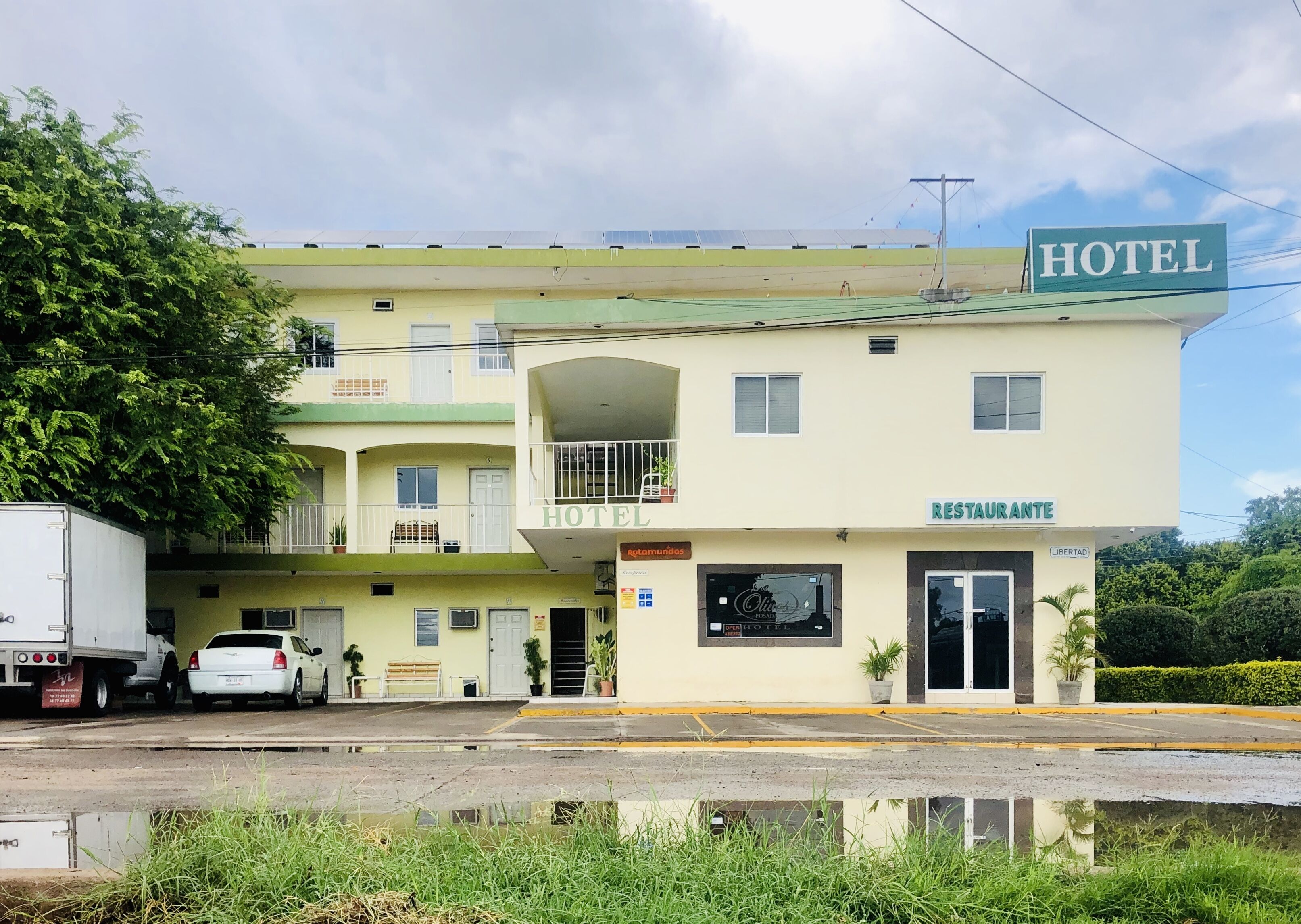 Edifício de dois andares com uma placa indicando que é um hotel e restaurante. Há carros estacionados e uma caminhonete na frente, e uma árvore é visível à esquerda.