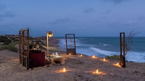 Montagem para refeições à beira-mar, com mesa, cadeiras e velas, com vista para o oceano.