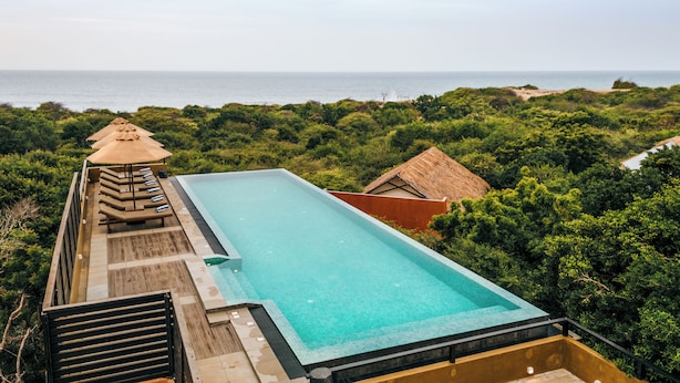 Uma piscina infinita no terraço, com espreguiçadeiras de madeira e uma estrutura com telhado de palha.