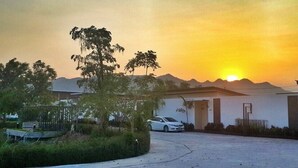 Uma área residencial com um carro branco estacionado em frente a uma casa, cercada por vegetação e um céu claro com o sol se pondo ao fundo.
