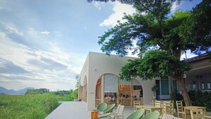 Uma área externa com mesas e cadeiras, entrada de um café e vista para a paisagem.