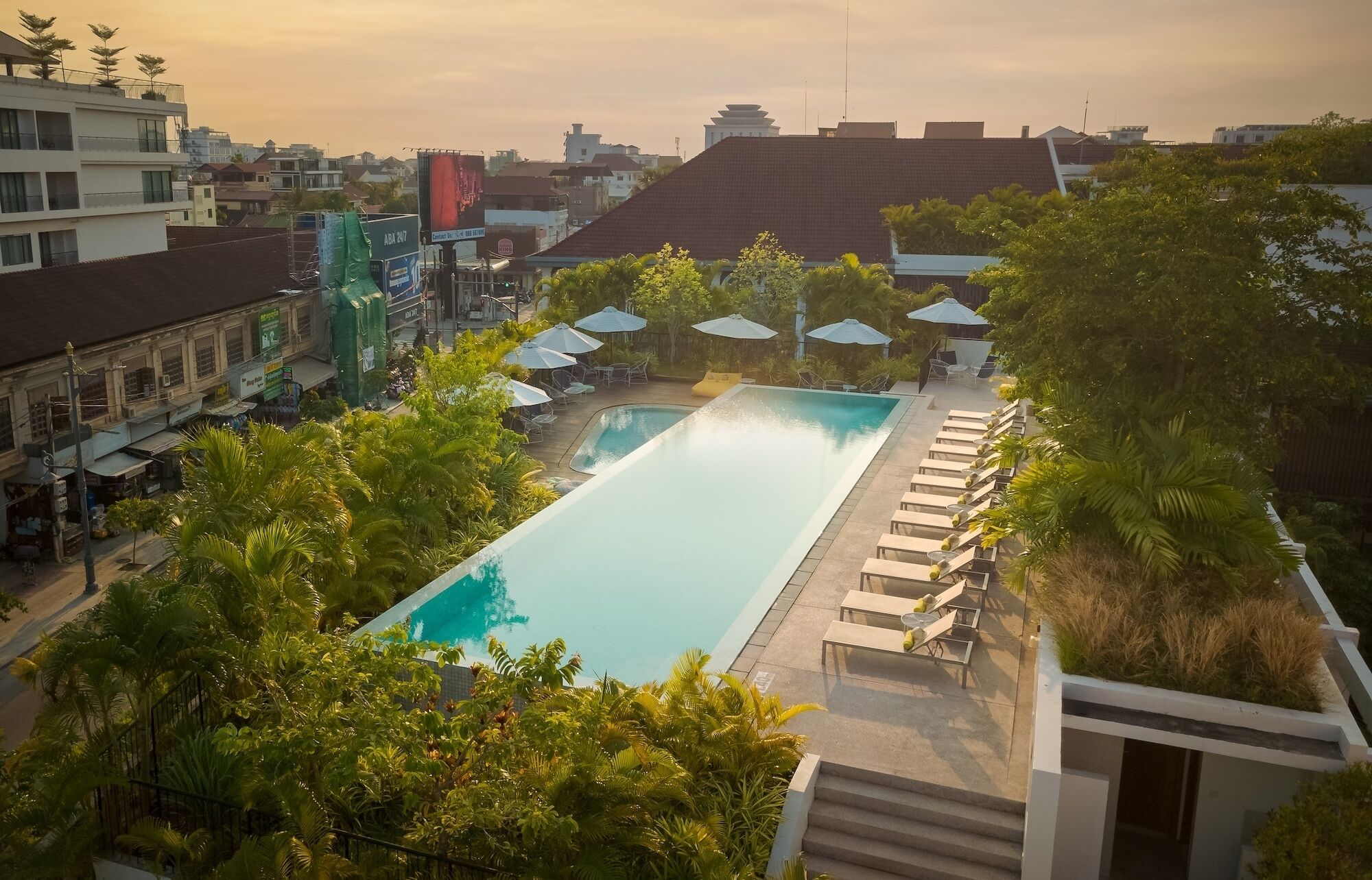 Piscina no terraço com espreguiçadeiras e guarda-sóis, cercada por vegetação e edifícios urbanos.