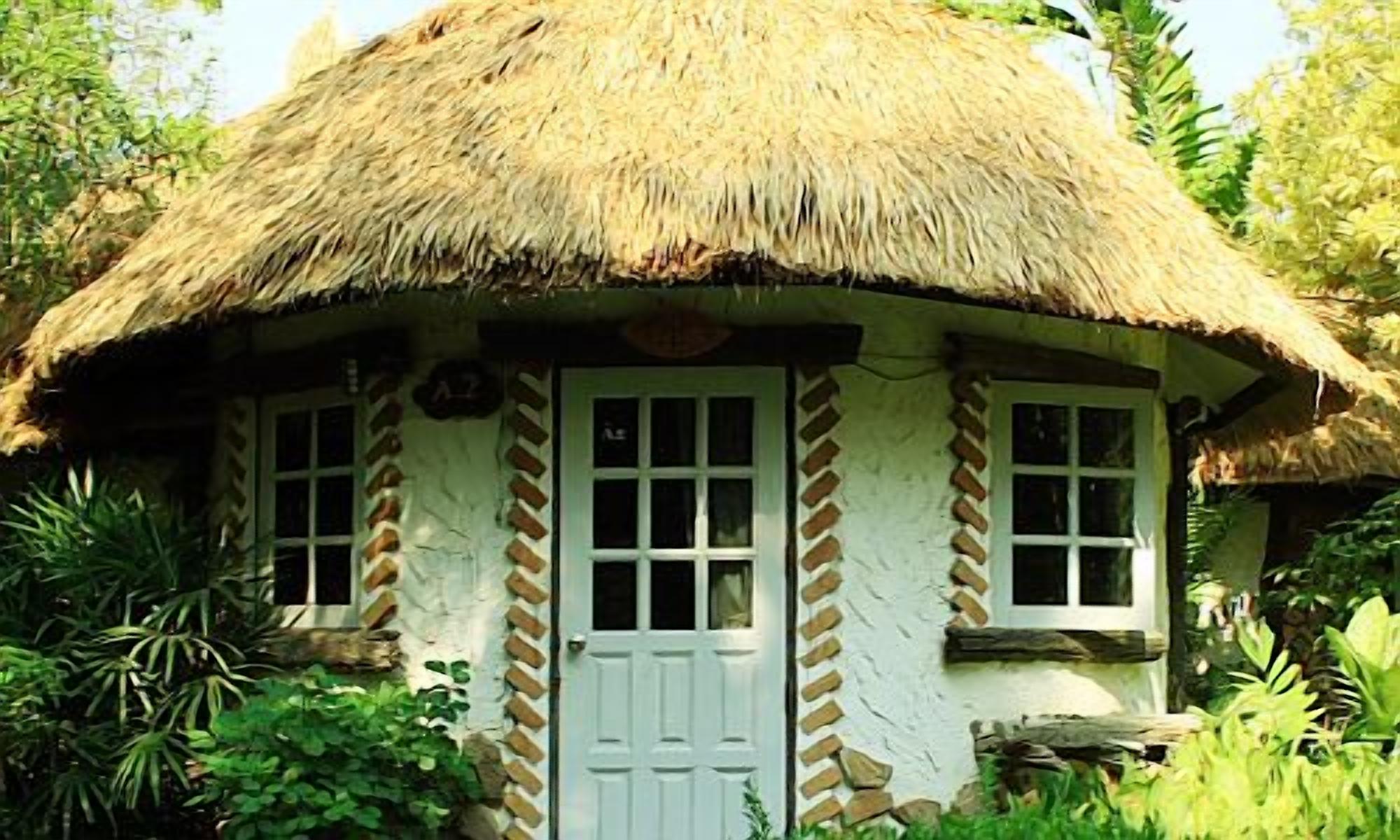 Uma cabana com telhado de palha, porta e janelas brancas, cercada por vegetação.