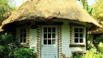 Uma cabana com telhado de palha, porta e janelas brancas, cercada por vegetação.