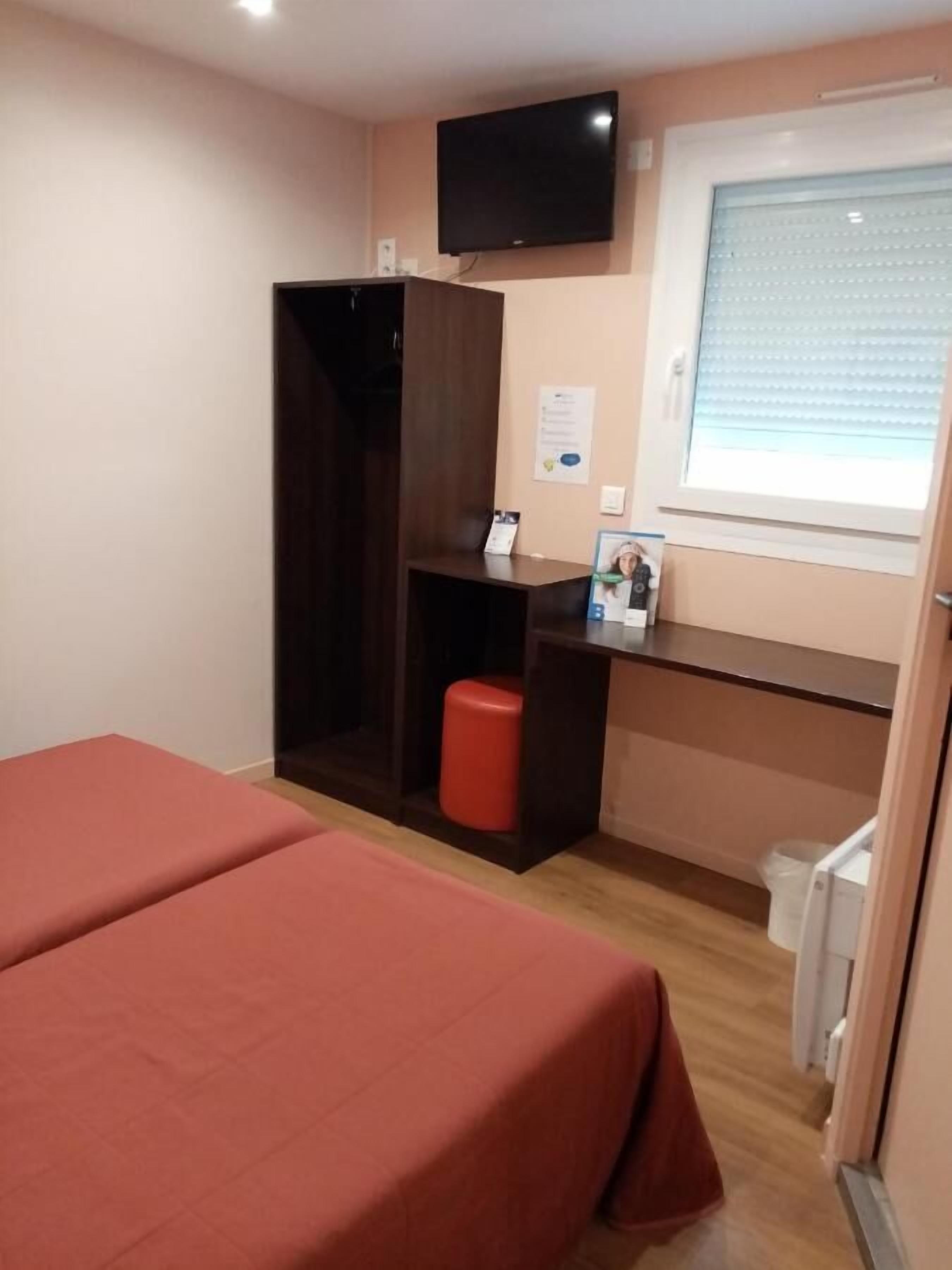 Quarto de hotel com duas camas, televisão presa na parede, janela com persianas e uma pequena mesa de trabalho com cadeira.