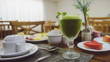 Uma mesa posta com uma bebida verde num copo, chávenas brancas, um prato com melancia e um vaso com flores roxas.