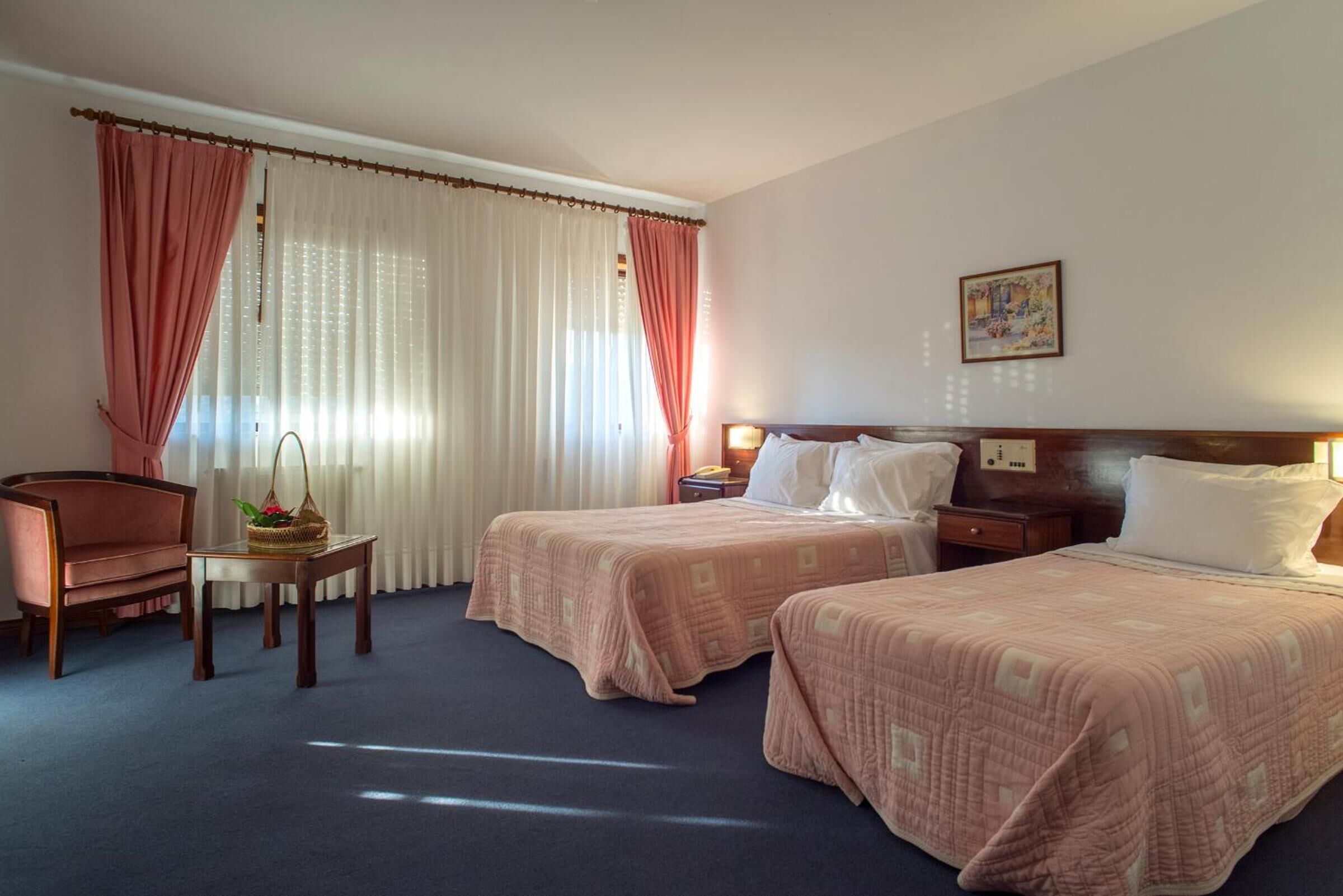Quarto de hotel com duas camas, uma cadeira, uma mesinha e um criado-mudo.