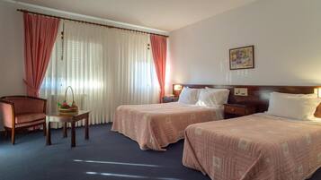 Quarto de hotel com duas camas, uma cadeira, uma mesinha e um criado-mudo.