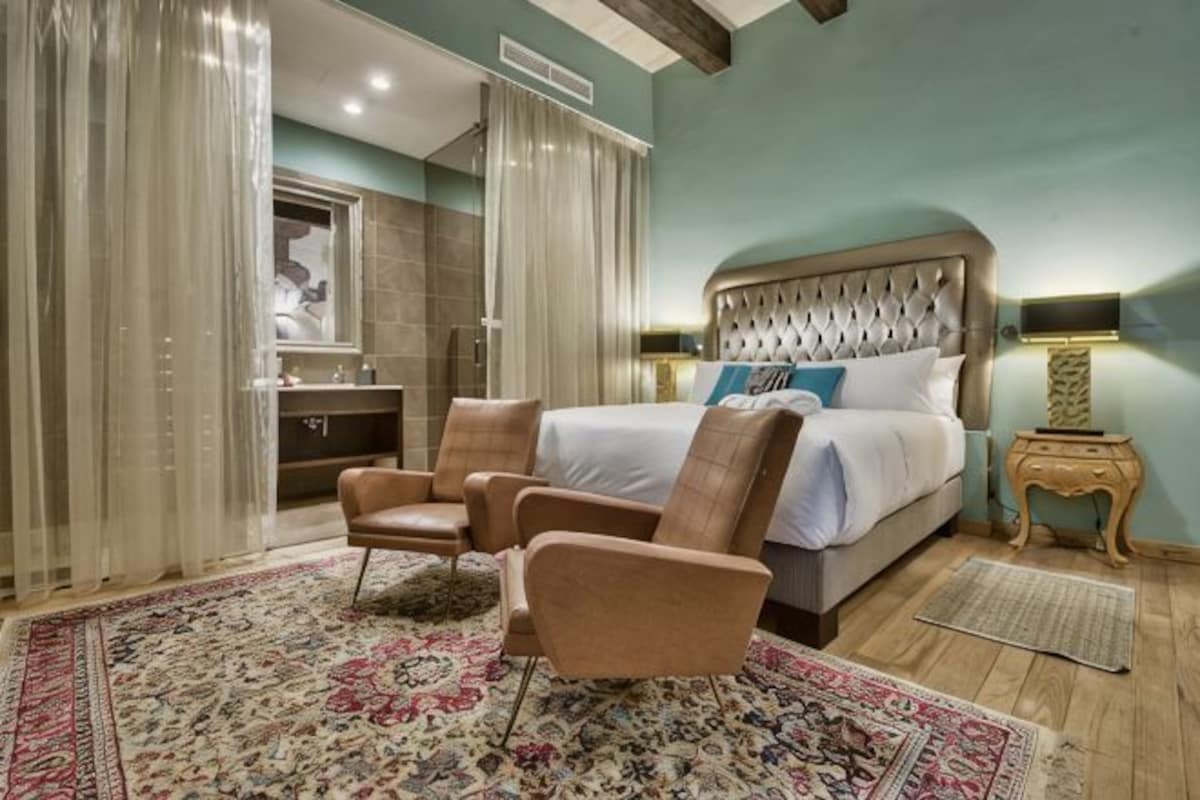 Quarto com cama, duas poltronas, mesa de cabeceira, espelho e banheiro com banheira, além de um tapete grande.