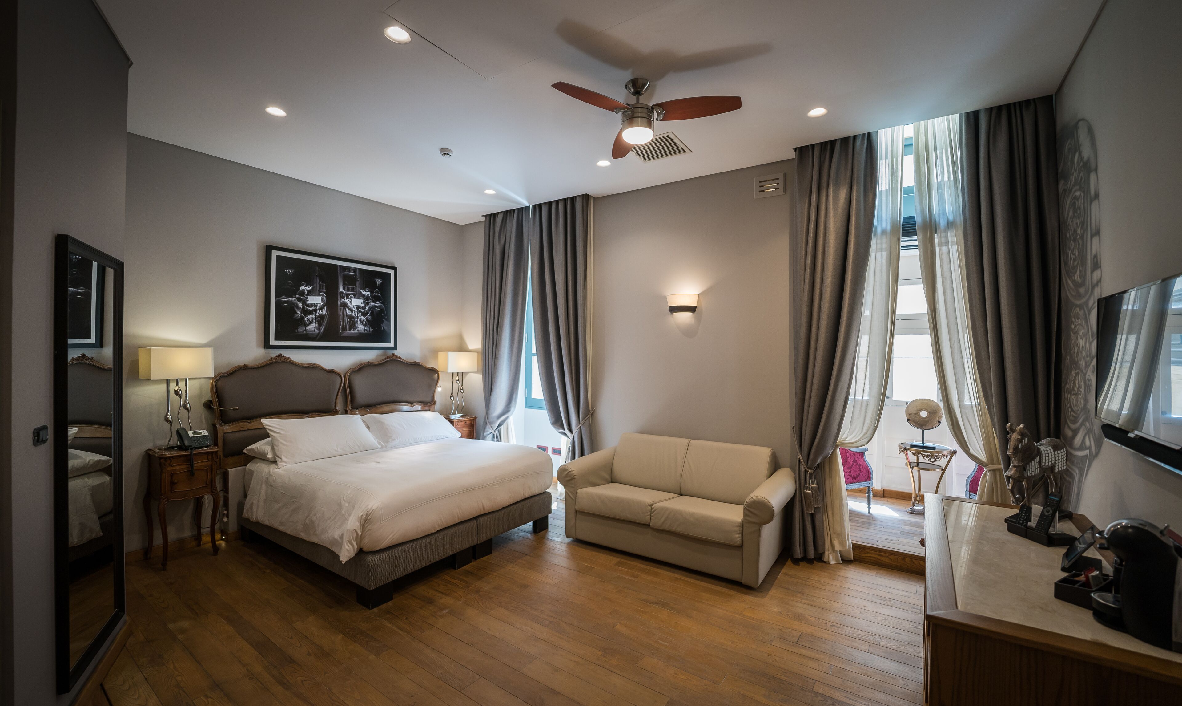 Quarto de hotel com cama, sofá, ventoinha no teto, televisão e janela com cortinas.