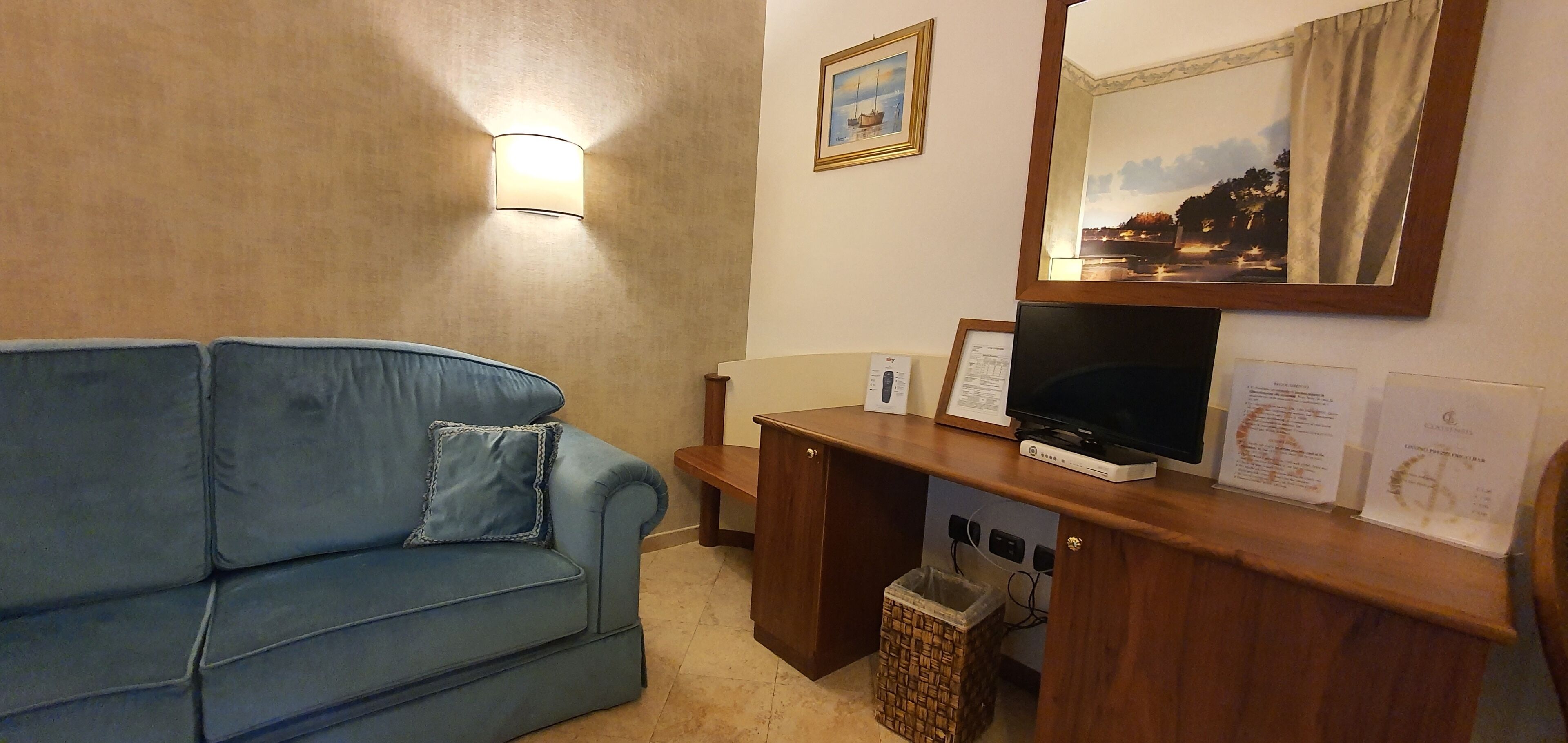 Quarto com sofá azul, escrivaninha de madeira com televisão, quadro emoldurado e uma lixeira pequena.