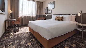 Quarto de hotel com uma cama grande, uma escrivaninha com um laptop, uma cadeira, uma mesa pequena com um abajur e uma janela com cortinas.