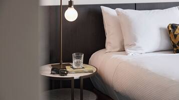 Uma cama bem arrumada com roupa de cama branca, uma mesa de cabeceira redonda com um copo e uma luminária.