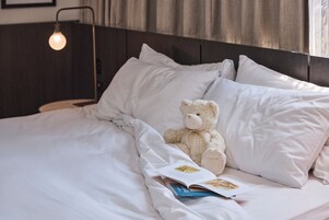 Cama bem arrumada com lençóis e almofadas brancas, um ursinho de pelúcia e um livro aberto sobre a cama.