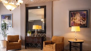 Um saguão de hotel com duas poltronas, uma mesinha de centro com abajur, um vaso de plantas, um espelho decorativo e uma pintura abstrata emoldurada.