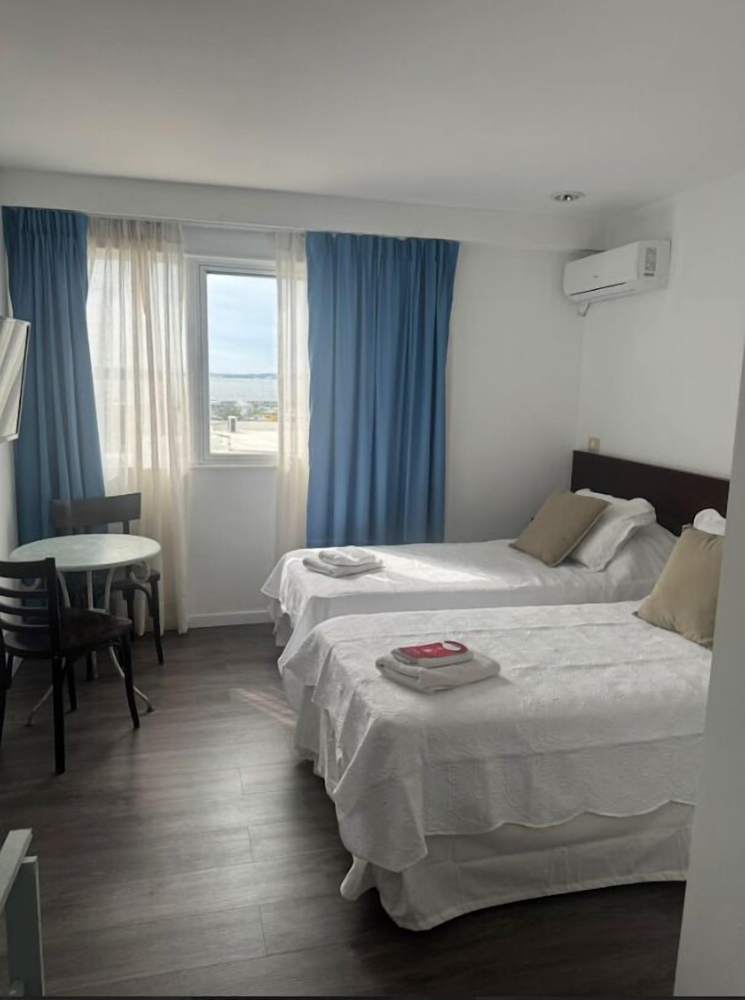Quarto de hotel com duas camas, uma mesa de jantar com cadeiras, janela com cortinas e aparelho de ar condicionado.