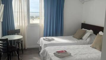 Quarto de hotel com duas camas, uma mesa de jantar com cadeiras, janela com cortinas e aparelho de ar condicionado.