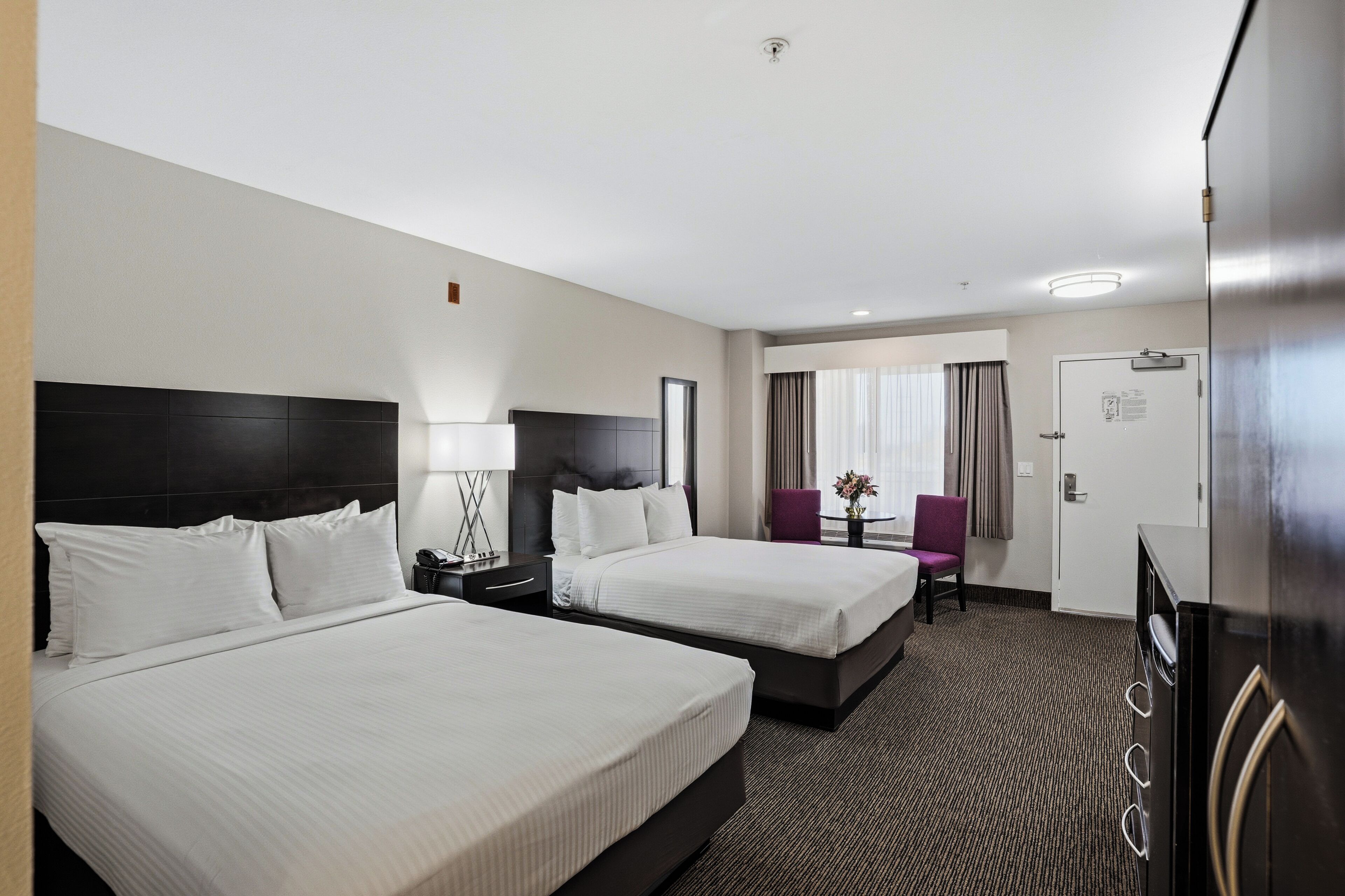 Quarto de hotel com duas camas, uma escrivaninha, uma cadeira, um frigorífico e uma porta com um aviso.