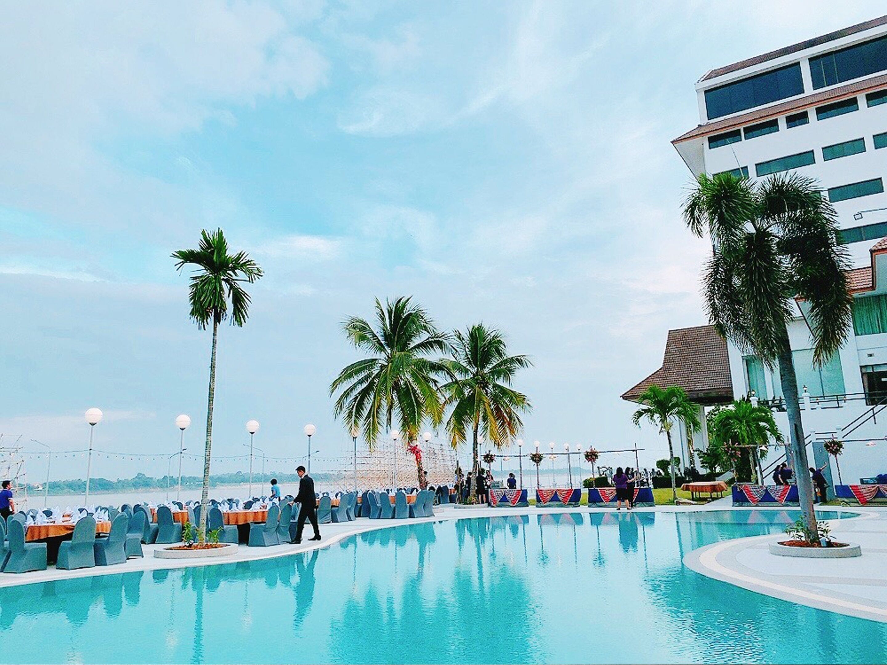 Piscina com água azul cristalina, rodeada por palmeiras e um edifício com vários andares.