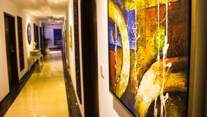 Corredor de hotel com obras de arte emolduradas nas paredes, piso polido e uma porta no final.