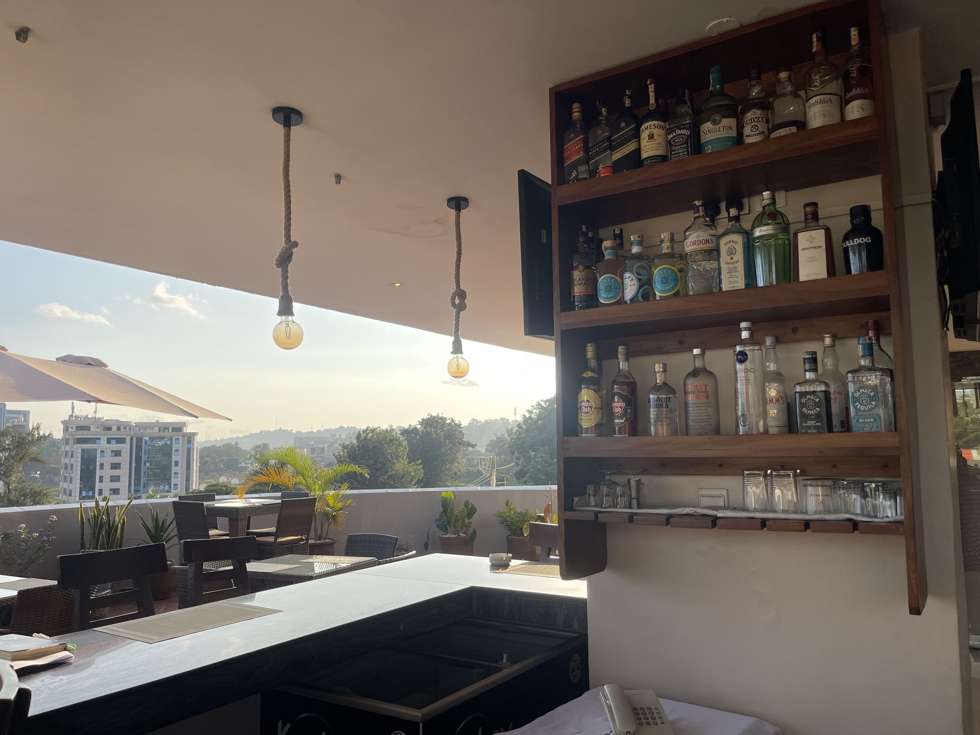 Um bar no terraço com diversas garrafas de bebidas em prateleiras, lâmpadas penduradas e área de estar ao ar livre com mesas e cadeiras.
