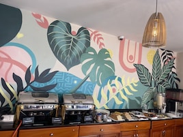 Um balcão de café da manhã com máquinas de café, diversas opções de comida e um mural colorido com folhas tropicais e formas abstratas.