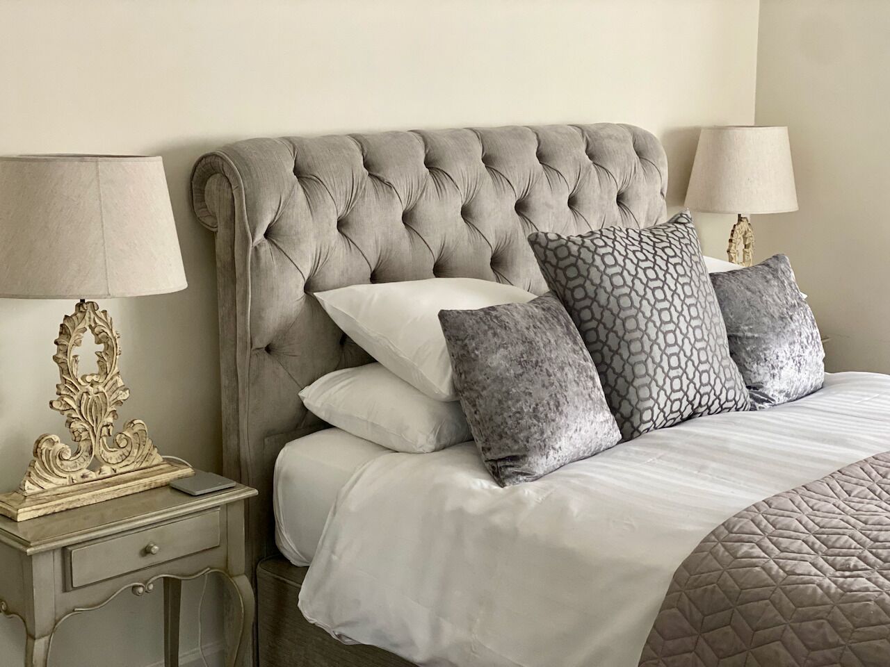 Cama bem arrumada com cabeceira estofada, roupa de cama branca e almofadas estampadas.