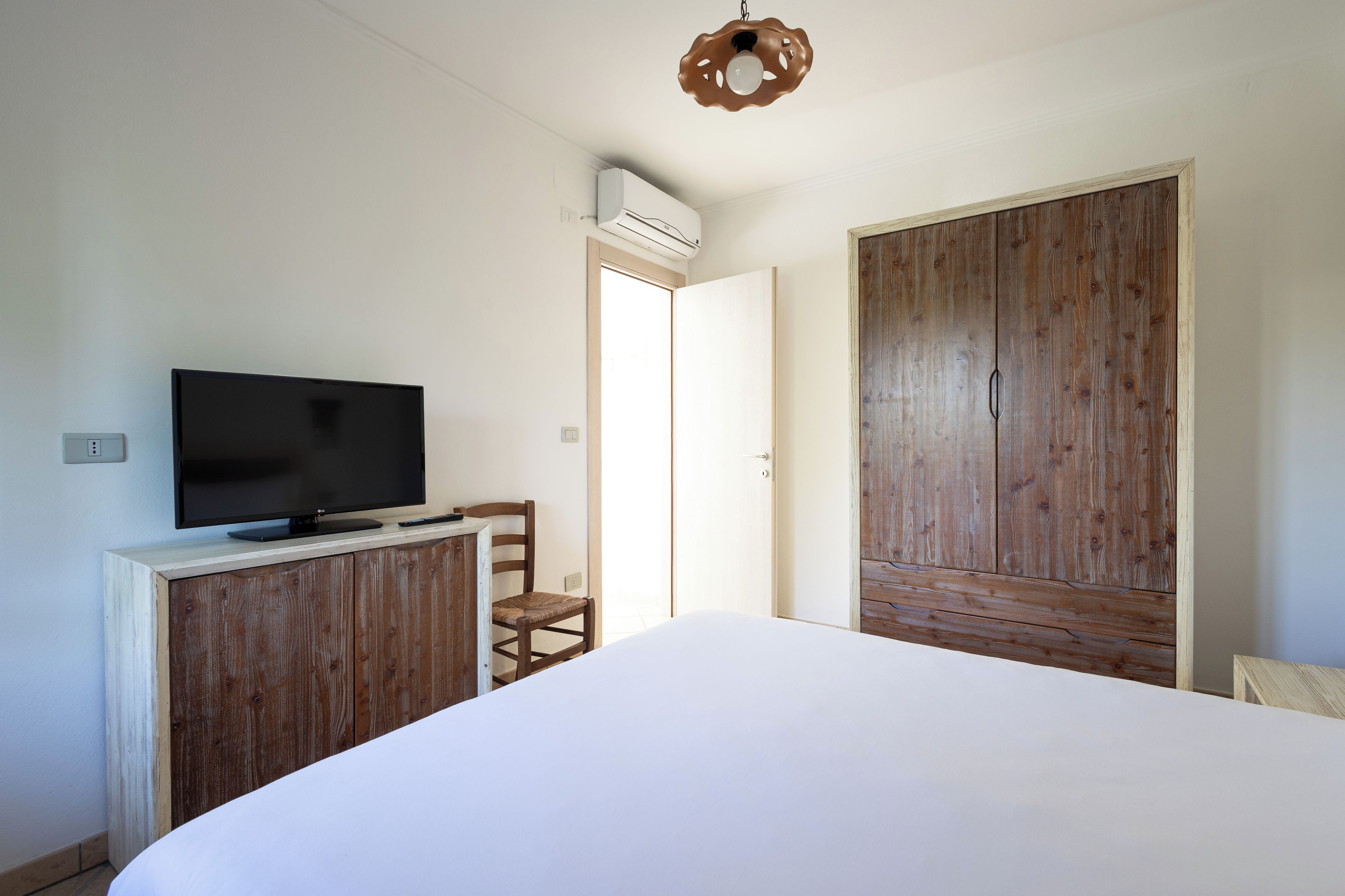 Quarto com cama, televisão sobre um armário de madeira, uma cadeira e um guarda-roupa.