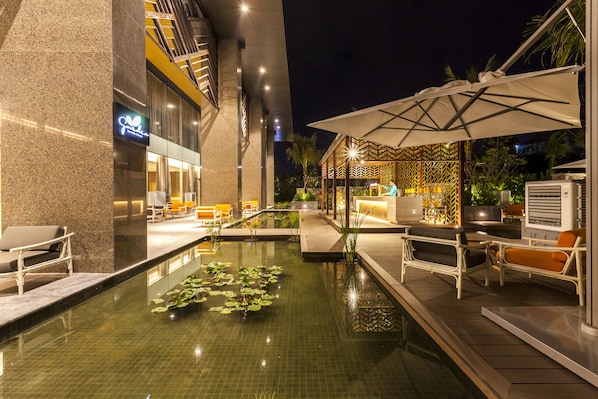 Uma área de piscina à noite, com espreguiçadeiras, um pequeno lago com vitórias-régias e um restaurante com mesas ao ar livre.