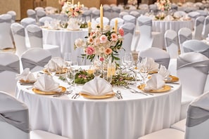 Uma sala de banquetes com mesas redondas dispostas para um evento formal, com toalhas brancas, talheres dourados e arranjos florais.