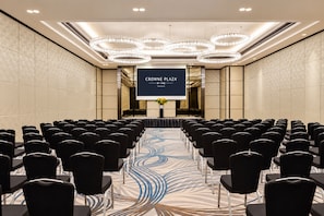 Uma sala de conferências com fileiras de cadeiras, um palco com uma tela exibindo 'CROWN PLAZA BY IHG' e um tapete estampado.