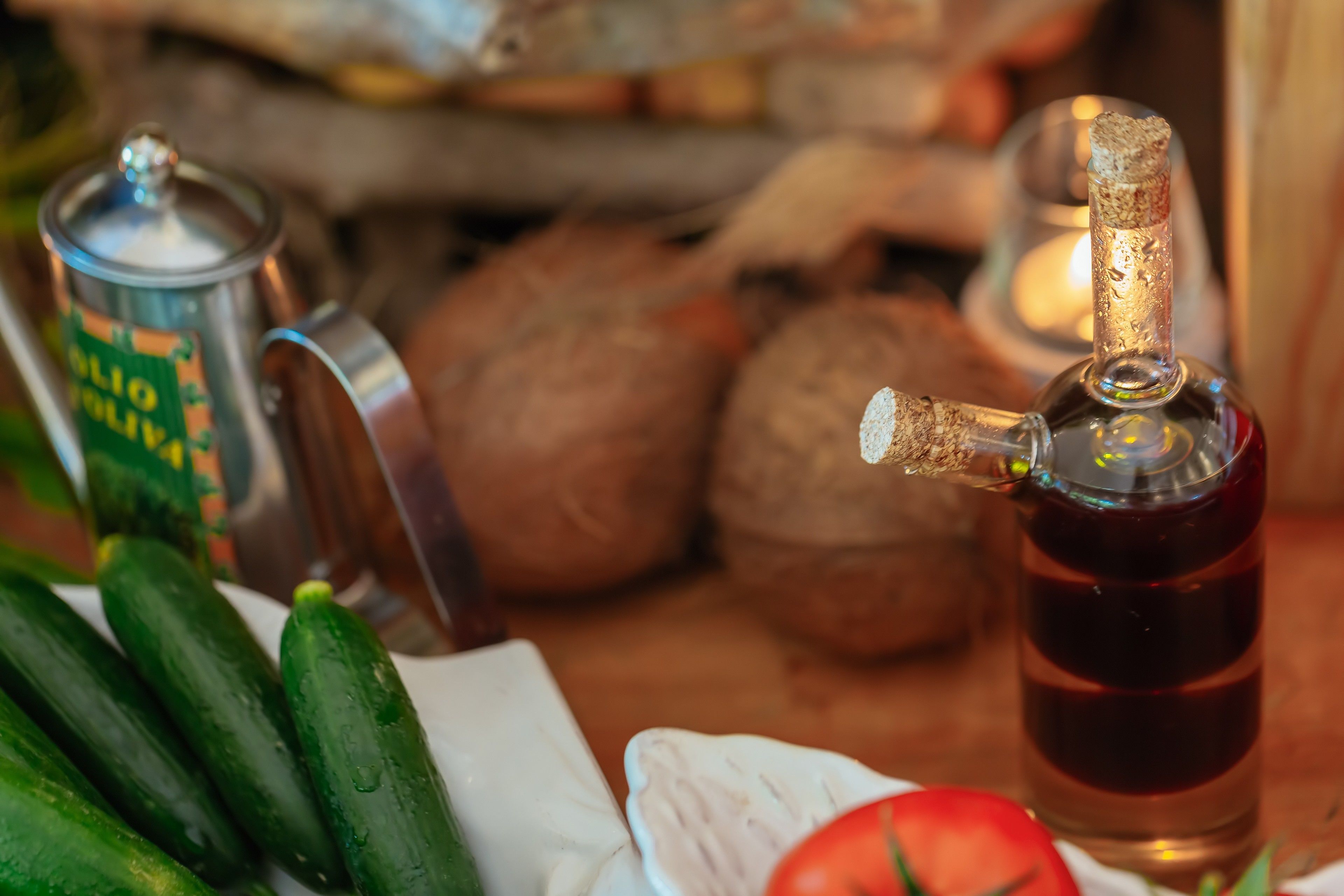 Uma garrafa com líquido escuro e rolha de cortiça, um recipiente de metal, pepinos, um tomate e um prato branco sobre uma superfície de madeira.