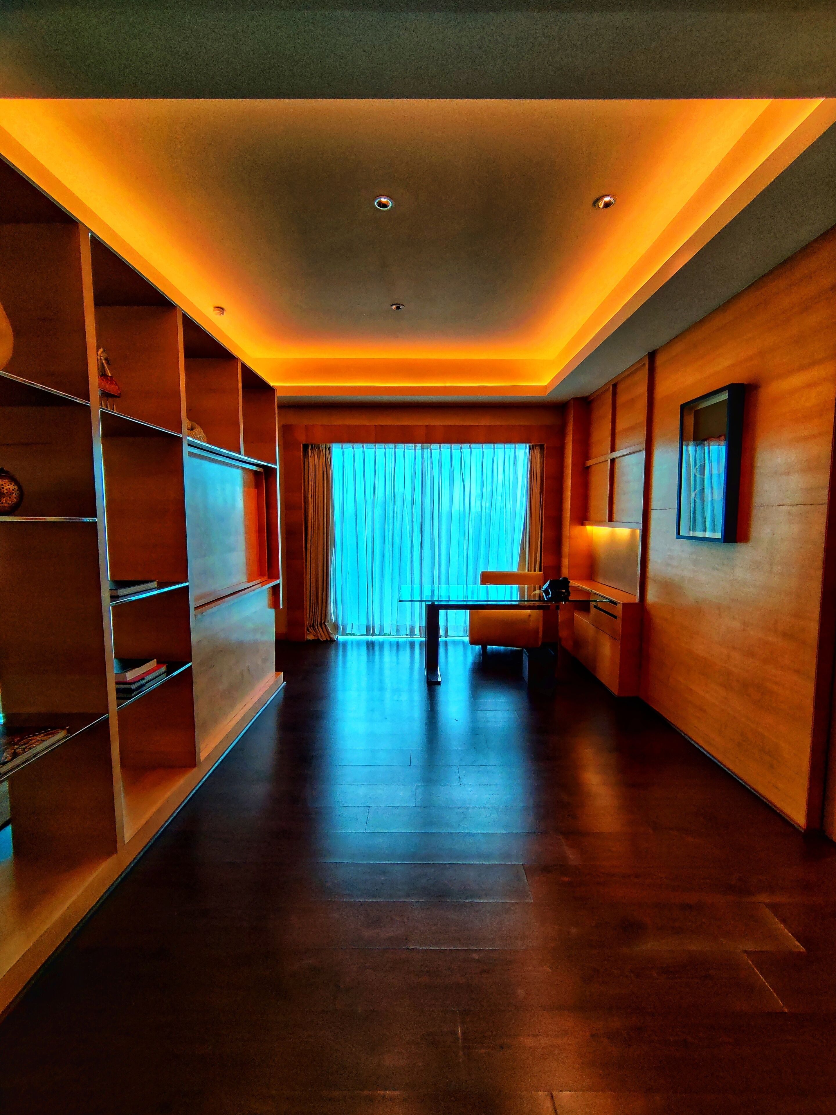 Corredor moderno de hotel com soalho de madeira, prateleiras embutidas, zona de estar e uma janela grande com cortinas azuis.